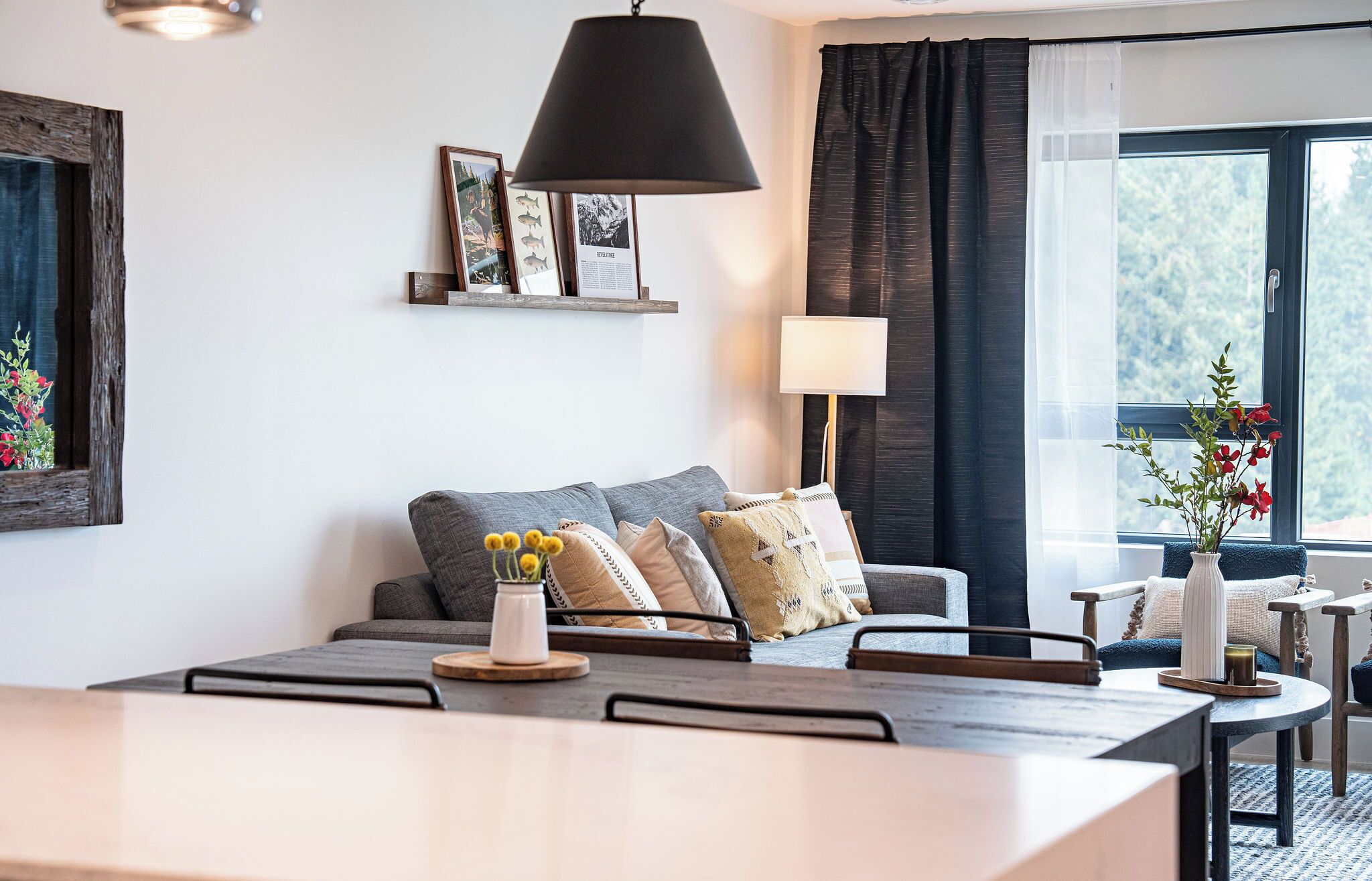 Uma sala de estar moderna com um sofá, uma mesa de centro e um espaço de jantar. Há quadros nas paredes, um abajur e uma janela com cortinas.