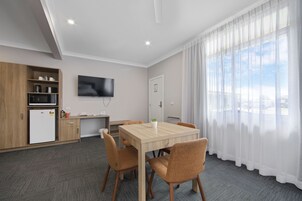 Quarto de hotel moderno com uma mesa de jantar de madeira, quatro cadeiras, televisão de tela plana na parede, um pequeno refrigerador e uma janela com cortinas leves.
