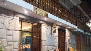 Entrada de hotel com paredes de pedra, uma varanda com balaústres de madeira e cadeiras coloridas do lado de fora.