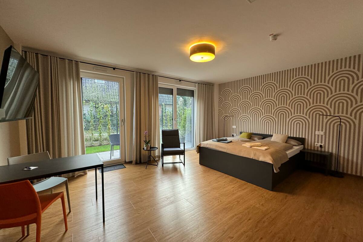 Quarto de hotel moderno com cama, escrivaninha e cadeira. Possui uma janela grande com cortinas, iluminação no teto e parede com padrão.
