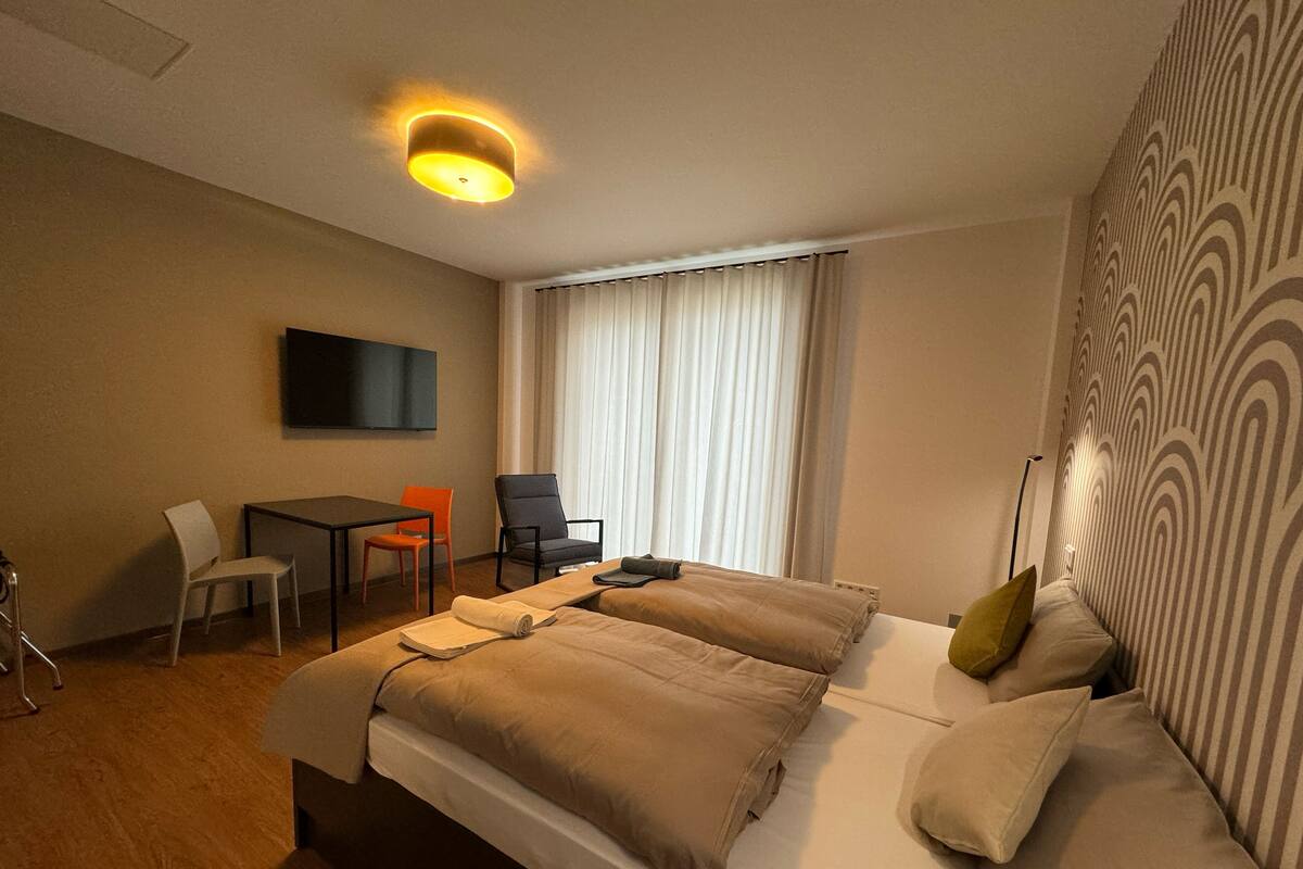 Quarto de hotel com duas camas, uma escrivaninha, uma cadeira, televisão e janela com cortinas.