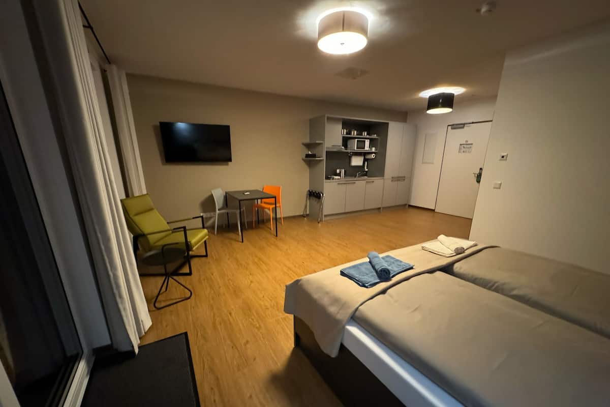 Quarto de hotel com cama, escrivaninha, cadeira, televisão e cozinha compacta.