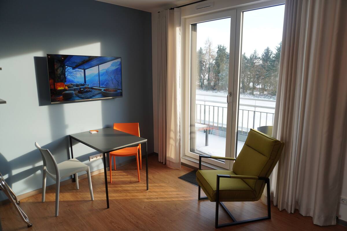 Quarto moderno com uma mesa de jantar, duas cadeiras, um sofá, um quadro na parede e uma janela grande com cortinas.