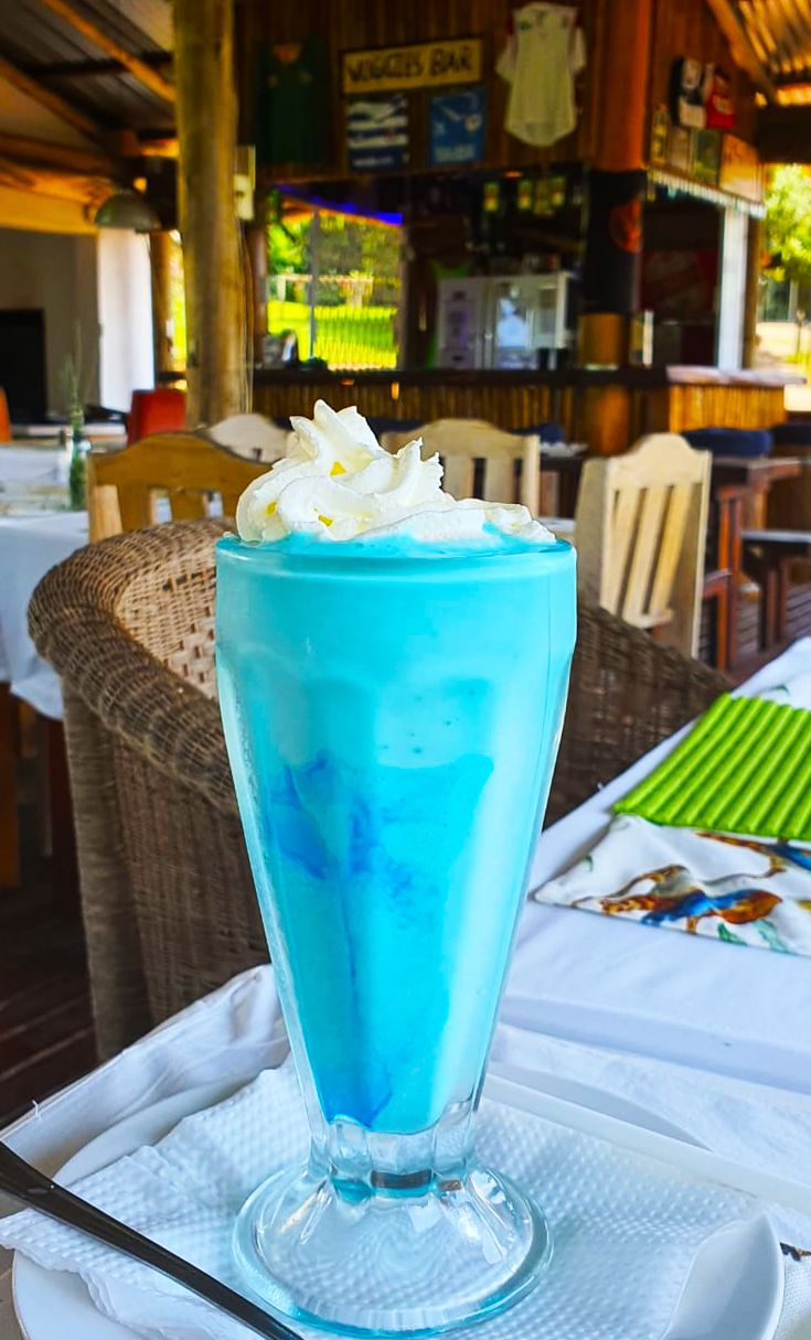 Um copo alto com bebida azul e cobertura branca, colocado sobre um guardanapo branco com uma colher ao lado.