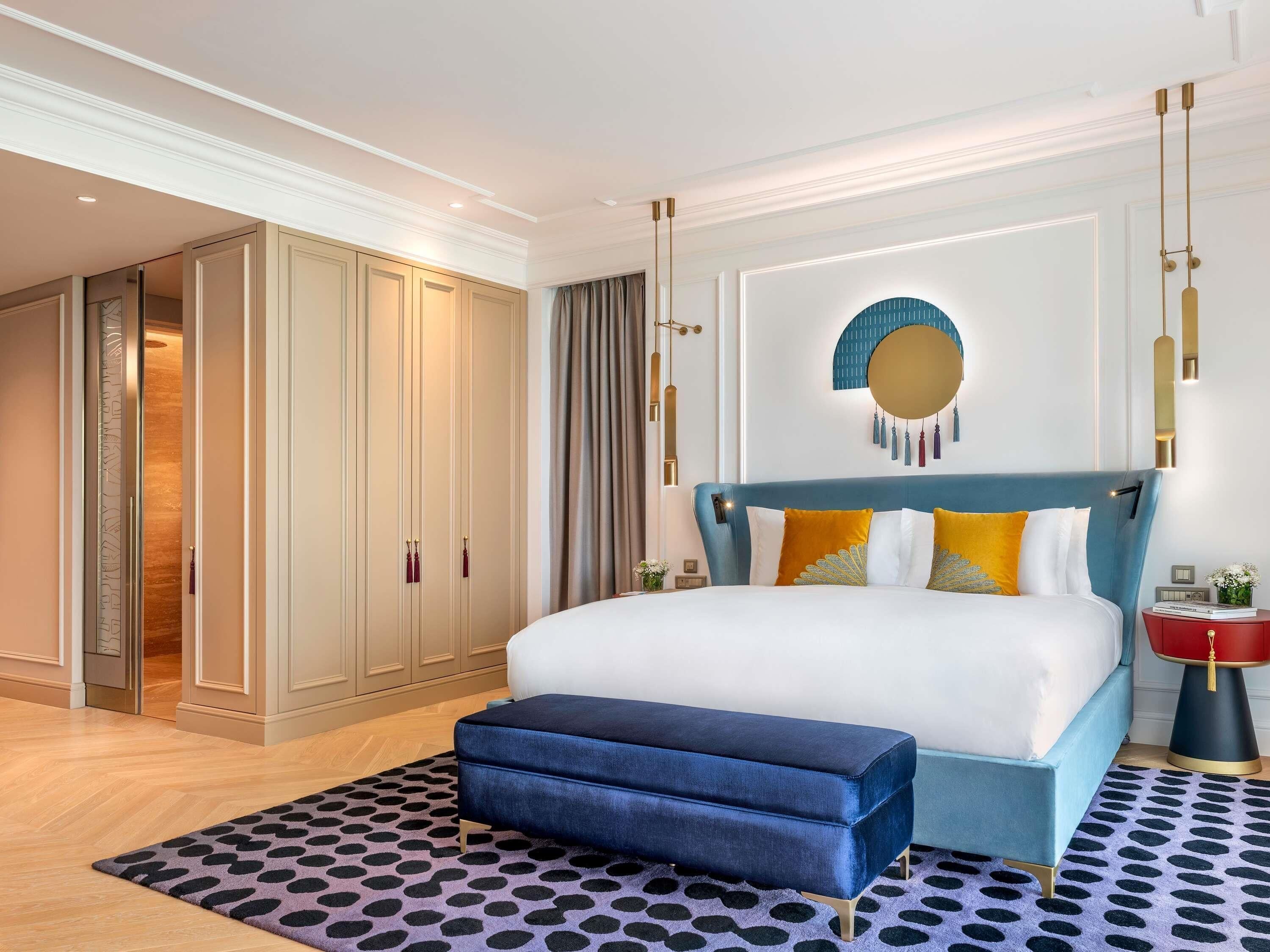 Quarto moderno com uma cama grande, um banquinho azul estofado, um espelho redondo e uma mesinha lateral vermelha.