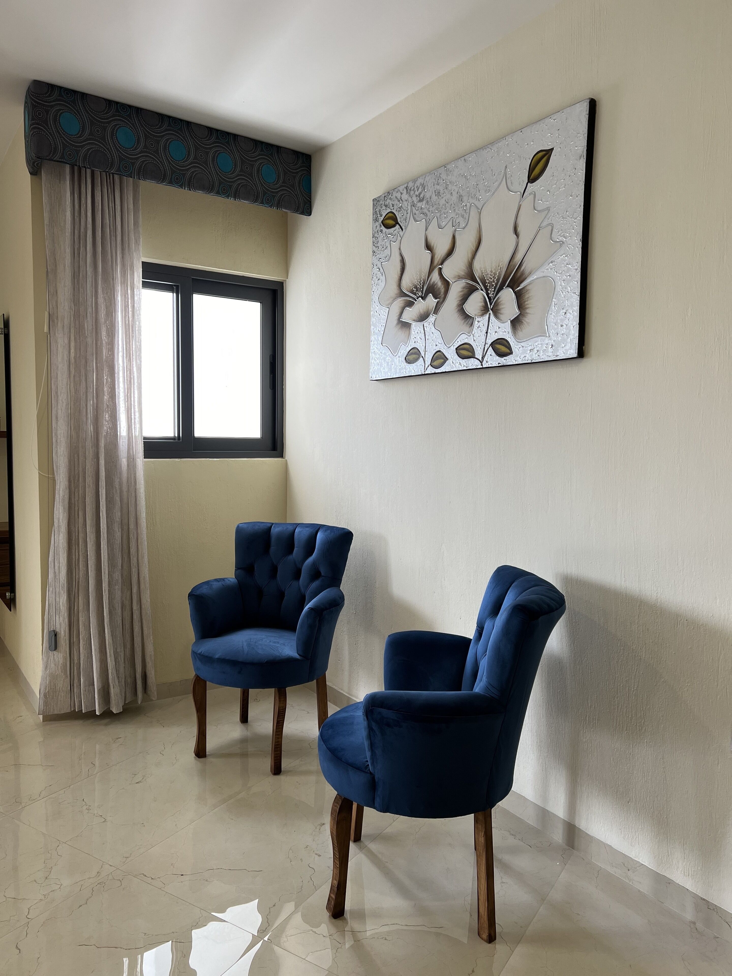 Quarto com duas poltronas azuis, uma janela com cortinas e uma obra de arte pendurada na parede.