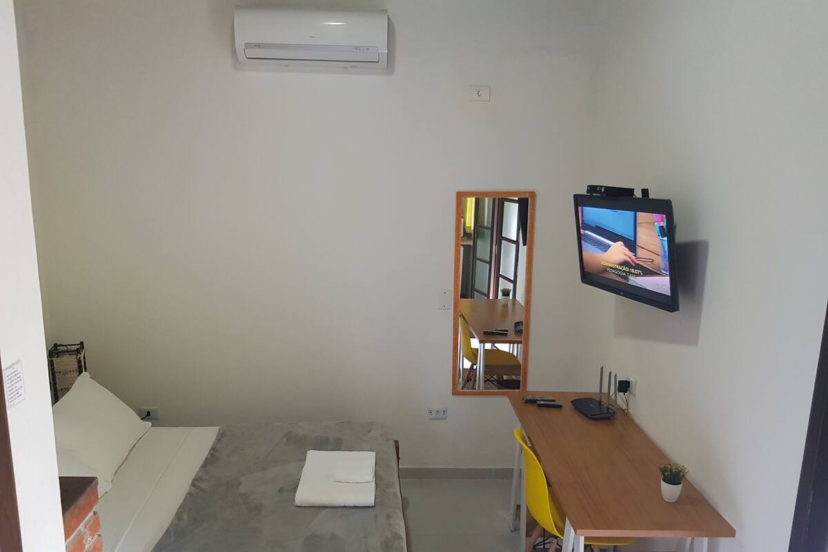 Quarto pequeno e limpo, com uma cama, escrivaninha, cadeira, televisão e espelho.