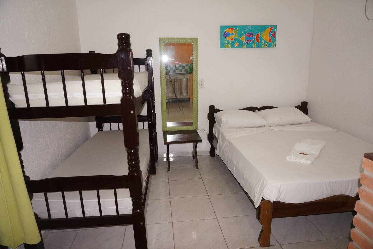 Quarto com duas camas de solteiro de madeira, uma cama individual, uma mesinha e um quadro de um peixe colorido na parede.