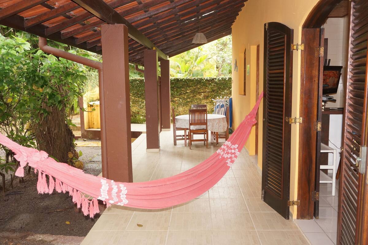 Uma área de varanda coberta com uma rede, uma mesa com cadeiras e vista para um jardim.