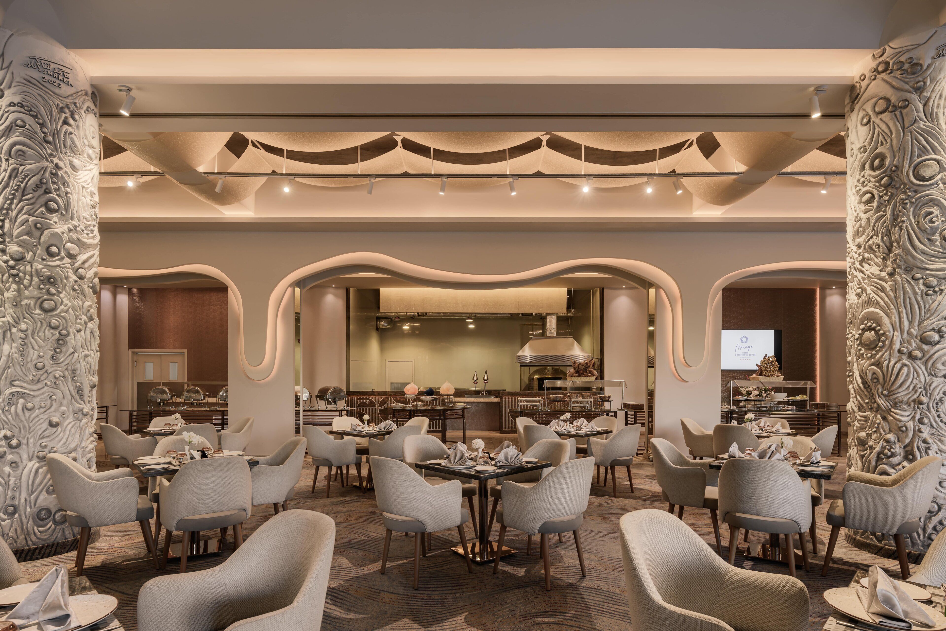 Um restaurante espaçoso e elegantemente decorado, com pé-direito alto, colunas ornamentadas e uma área de jantar central com mesas redondas e assentos confortáveis.