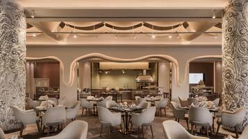 Um restaurante espaçoso e elegantemente decorado, com pé-direito alto, colunas ornamentadas e uma área de jantar central com mesas redondas e assentos confortáveis.