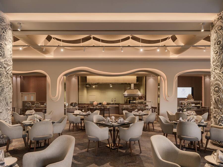Um restaurante espaçoso e elegantemente decorado, com pé-direito alto, colunas ornamentadas e uma área de jantar central com mesas redondas e assentos confortáveis.