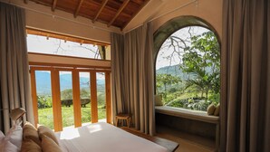 Quarto com cama grande, vista para áreas verdes e montanhas pela janela, e um banco embutido com almofadas.