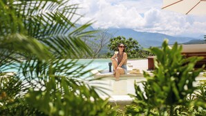 Uma pessoa sentada à beira da piscina, com vista para uma montanha ao fundo.