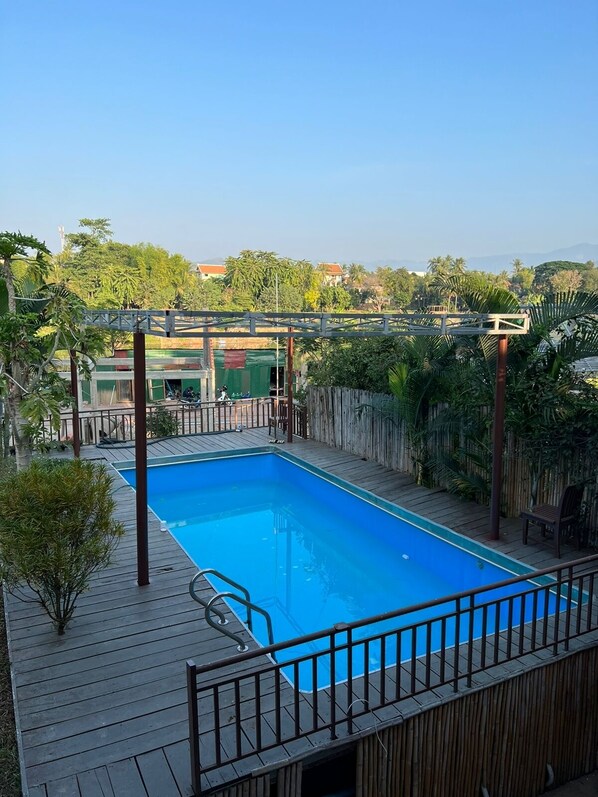 Piscina retangular com deck de madeira e guarda-corpos de metal, cercada por um cercado de bambu e diversas plantas.