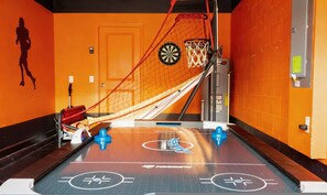 Mesa de bilhar com rede, alvo de dardos e cesta de basquete em um salão com paredes laranja.