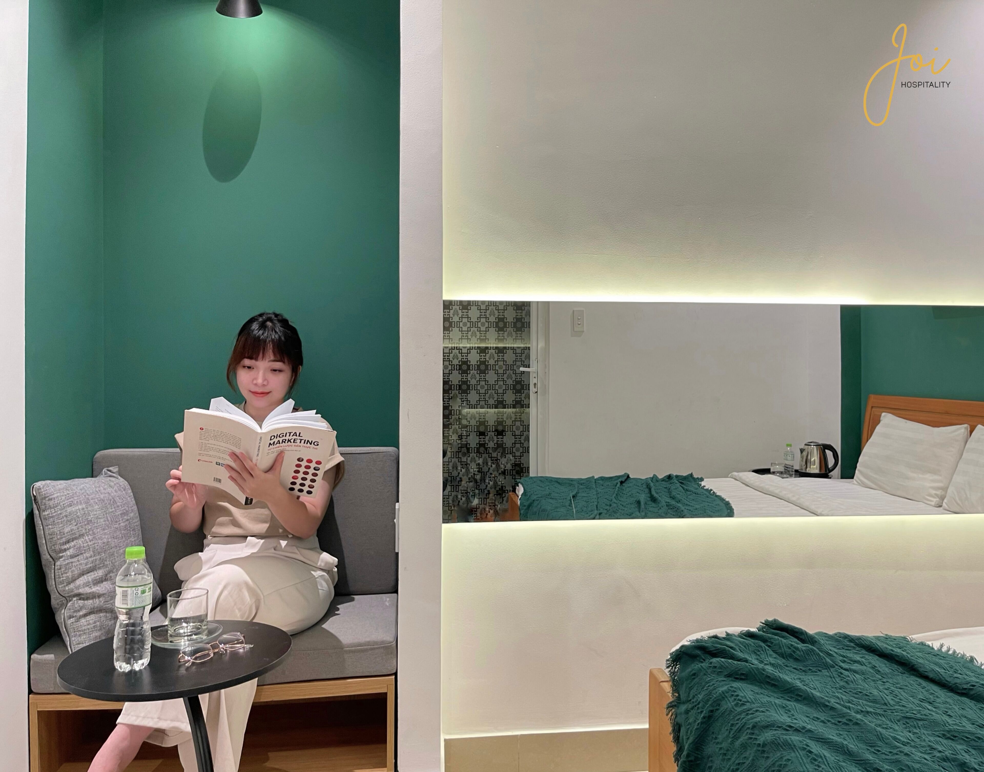 Uma pessoa lendo um livro em um quarto com parede verde, uma cama e uma mesinha com uma garrafa e copos.
