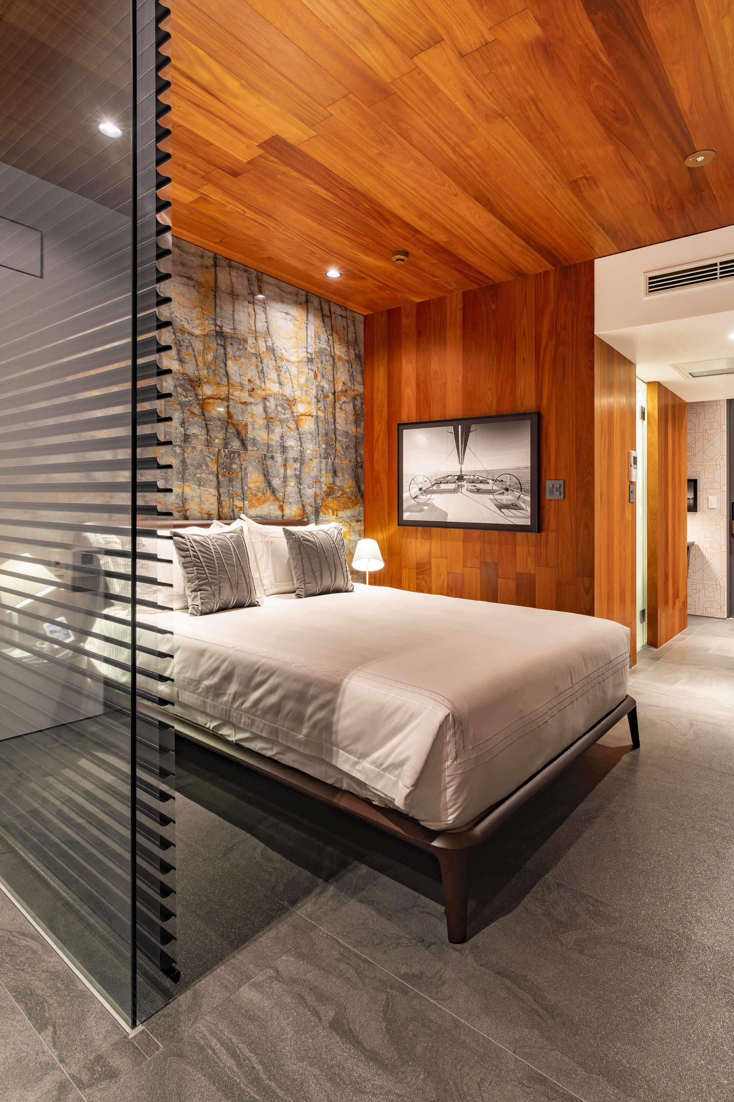 Quarto moderno com teto de madeira, uma cama grande com colchão branco e uma parede que imita mármore atrás da cama.