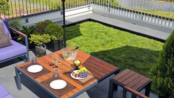 Um pátio externo com mesa posta para refeições, rodeado por assentos roxos, com vista para uma paisagem deslumbrante com montanhas e um rio.