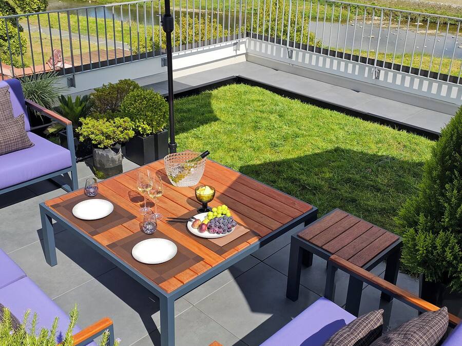 Um pátio externo com mesa posta para refeições, rodeado por assentos roxos, com vista para uma paisagem deslumbrante com montanhas e um rio.