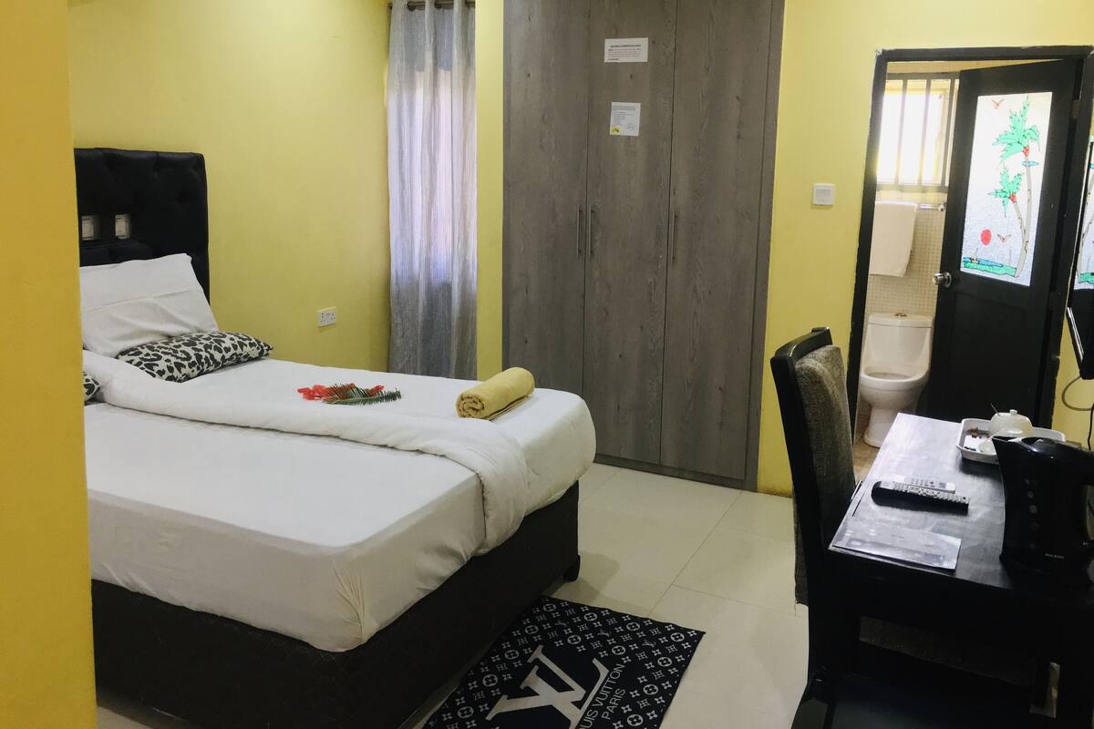 Quarto de hotel com cama, escrivaninha, cadeira, guarda-roupa e banheiro visível através de uma porta aberta.