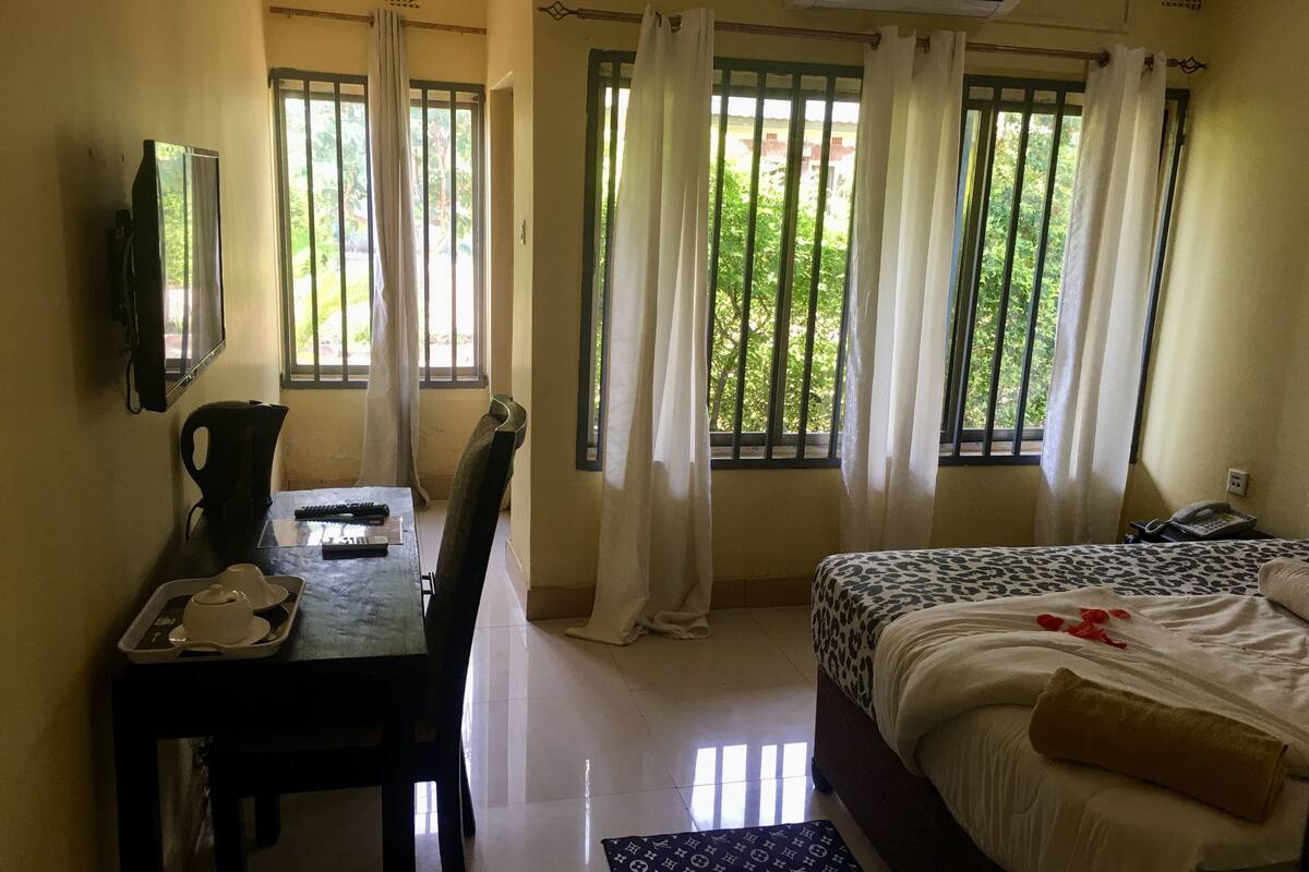 Quarto de hotel com cama, escrivaninha, telefone, cadeira e janela com cortinas.