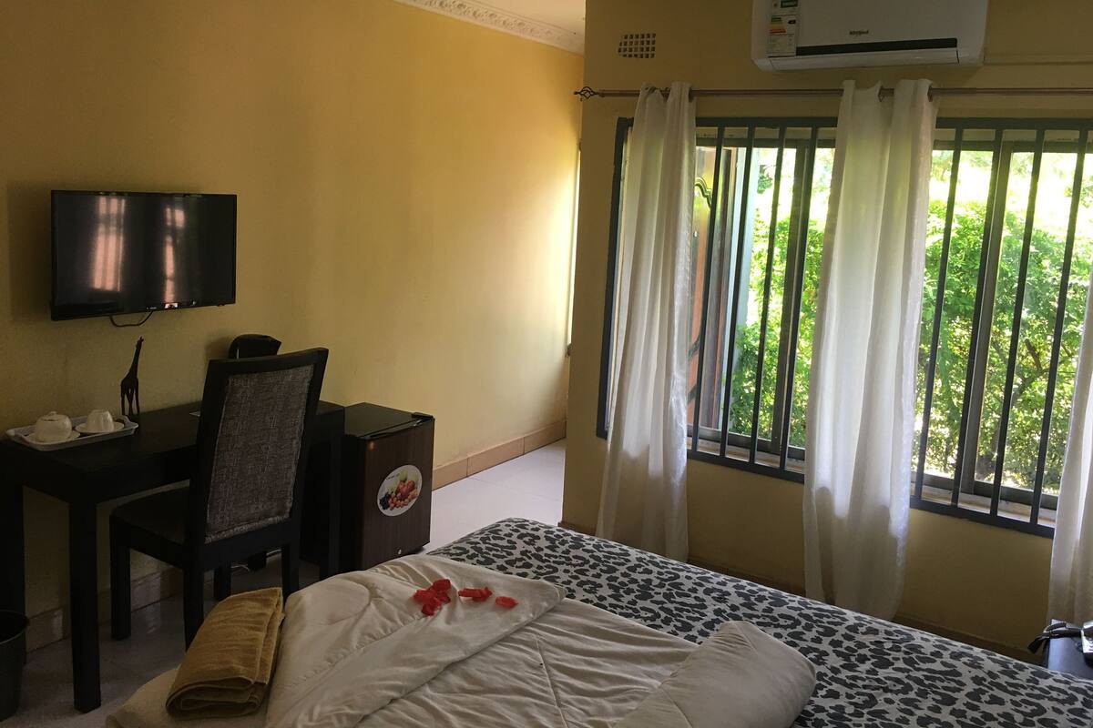 Quarto de hotel com cama, escrivaninha com cadeira, televisão e janela com cortinas.