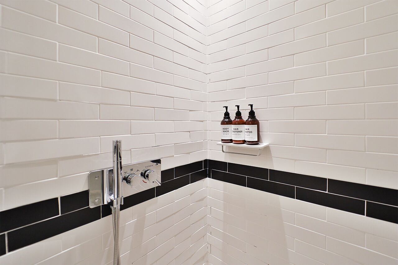 Casa de banho com azulejos brancos tipo metrô, uma moldura preta e branca, e um chuveiro com torneira cromada. Há três frascos de gel de banho e loção numa prateleira.
