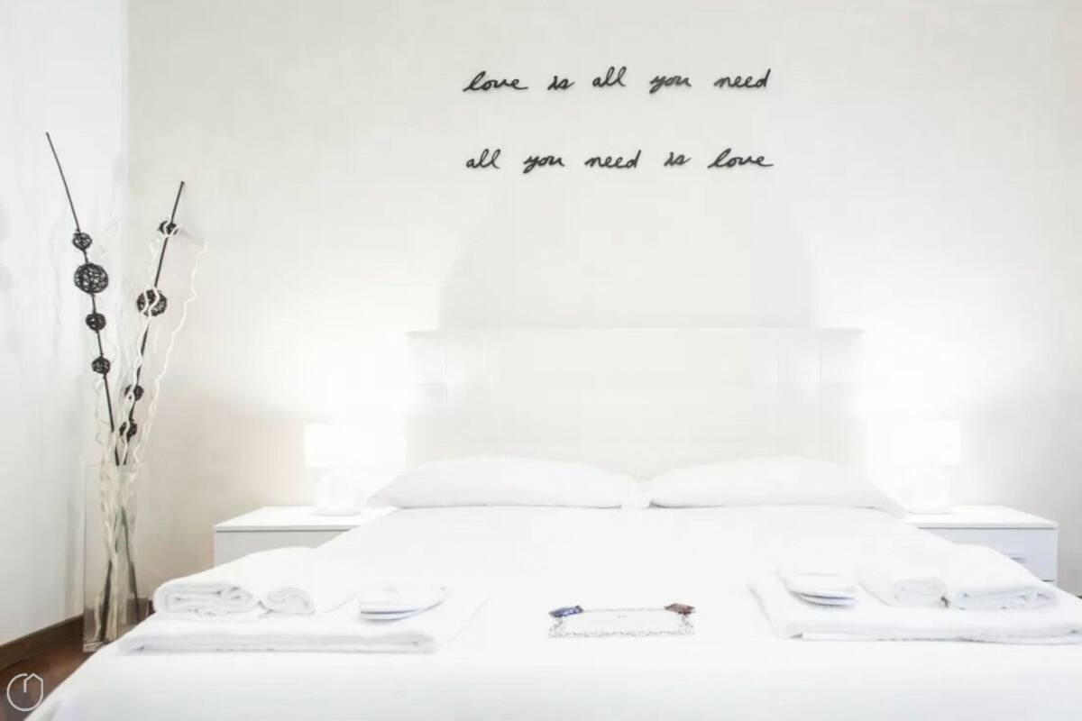 Uma cama bem arrumada com roupa de cama branca, uma mesinha de cabeceira com abajur e uma parede com uma frase motivacional.