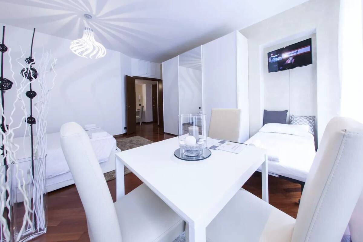 Um quarto moderno com decoração em branco, incluindo uma mesa de jantar, cadeiras, um sofá e uma TV fixada na parede.