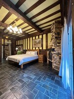 Quarto com lareira de pedra, cama com colcha branca e teto de madeira.