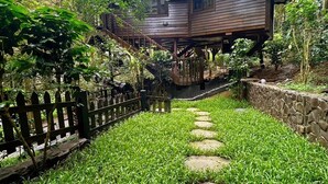 Casa de madeira construída sobre pilares, rodeada por vegetação exuberante e um muro de pedra.