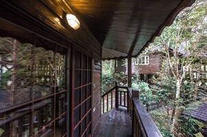 Um deck de madeira com guarda-corpo, grandes janelas e vista para a floresta ao redor.