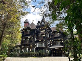 Um edifício grande e de vários andares com telhado íngreme, várias chaminés e uma combinação de lambris de madeira escura e clara.