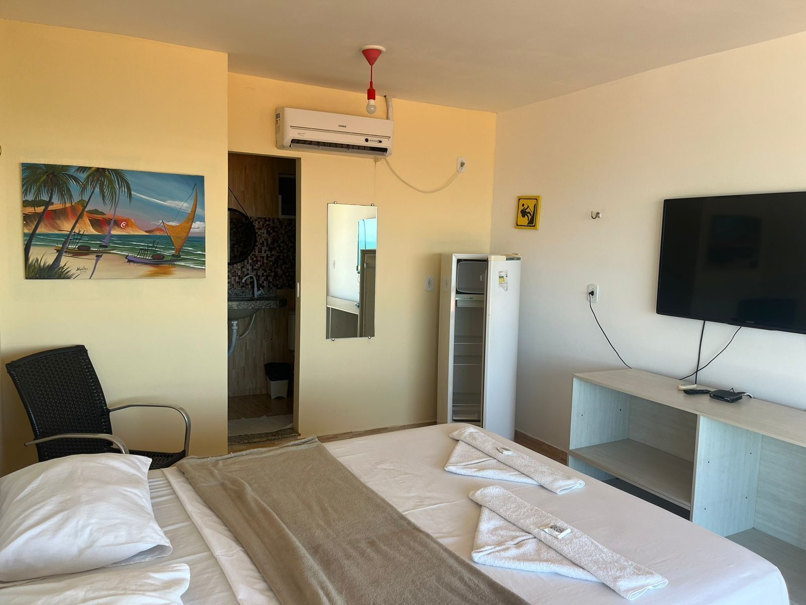 Quarto de hotel com cama, poltrona, televisão, ar condicionado e um quadro na parede.
