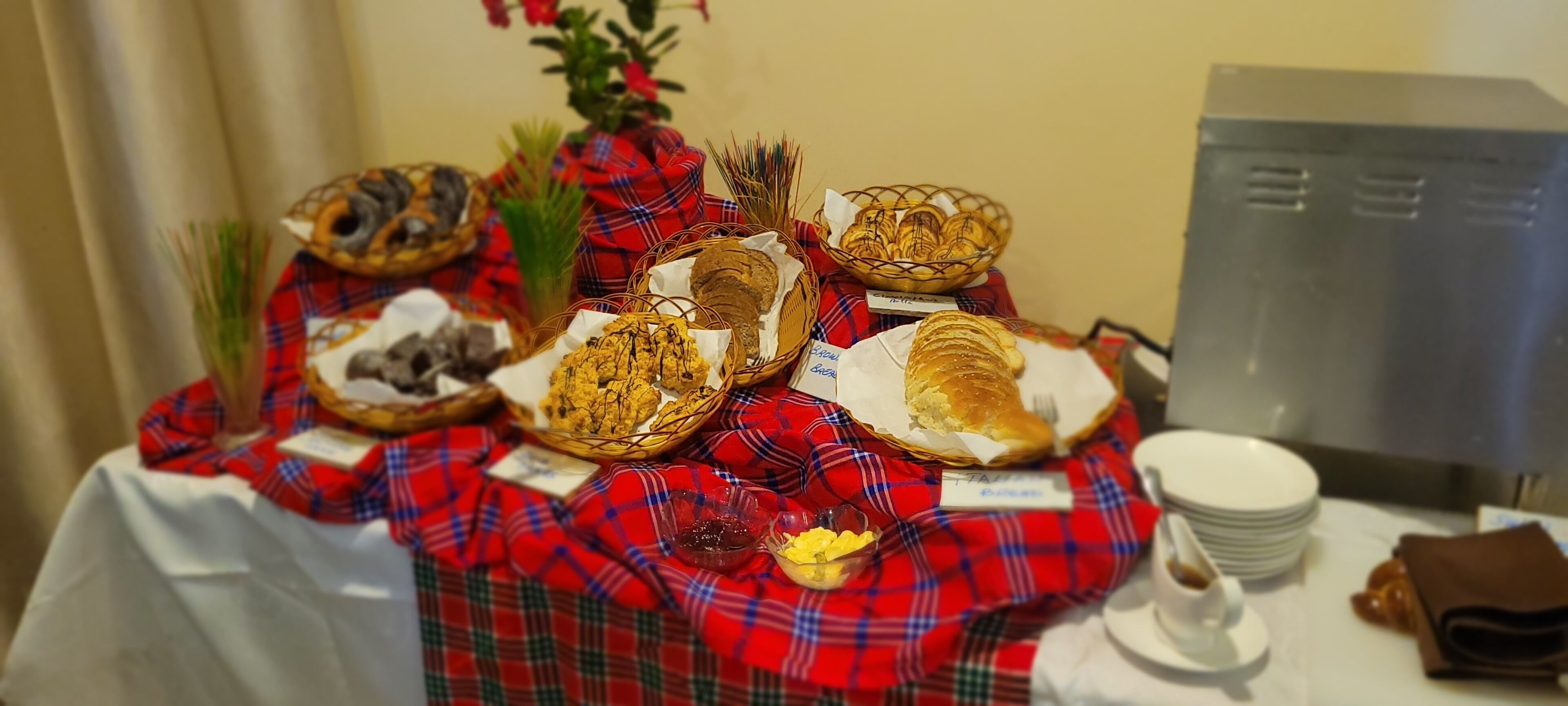 Uma mesa de buffet com diversos doces e uma estação de café, coberta por uma toalha xadrez vermelha e azul.
