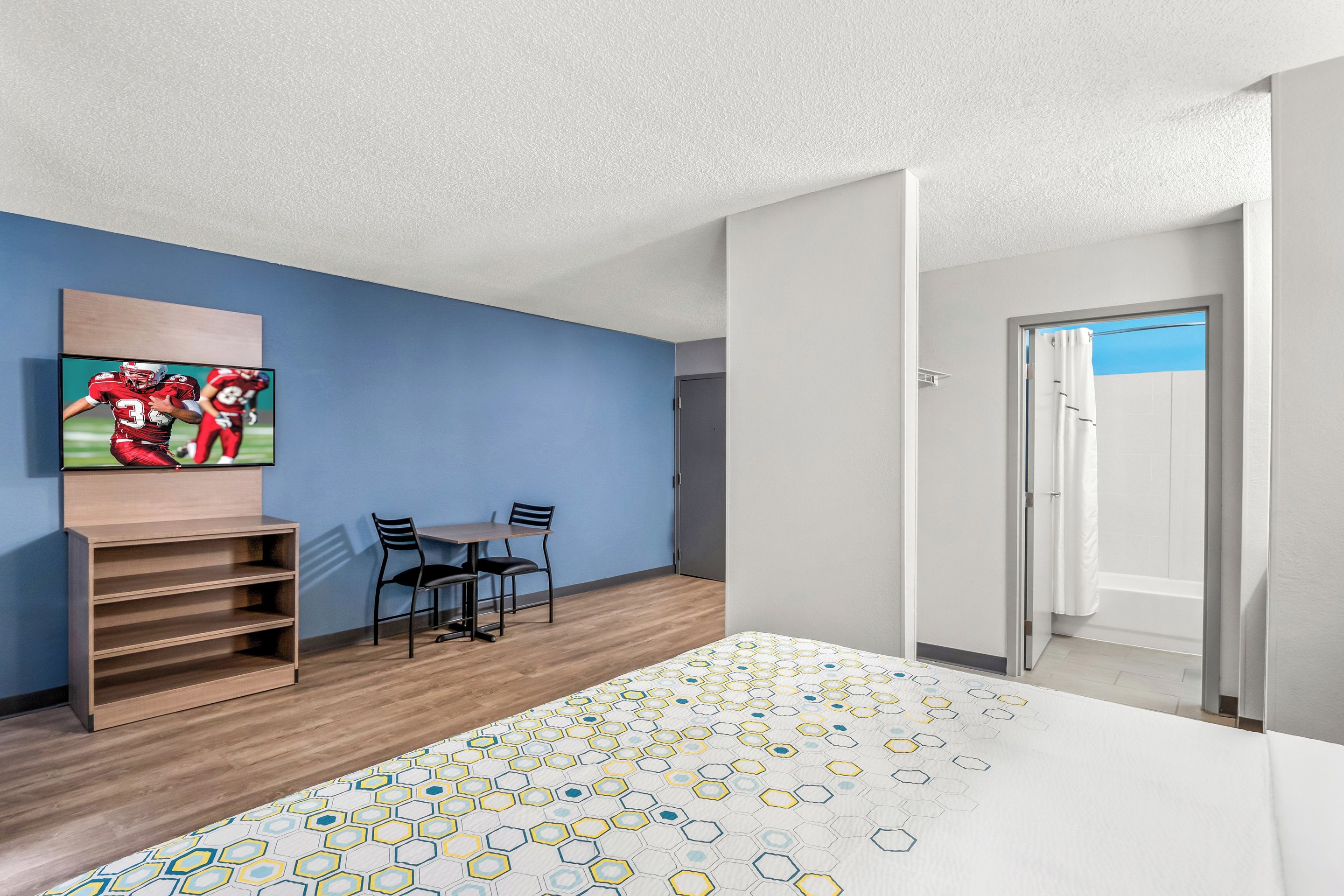 Quarto de hotel com cama, televisão na parede, secretária com cadeiras e zona de duche.
