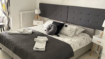 Cama bem arrumada com cabeceira cinza, roupa de cama branca e manta preta. Tem uma mesa de cabeceira com luminária, radiador e um quadro emoldurado na parede.