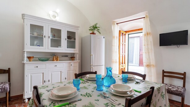 Uma sala de jantar com uma mesa posta para quatro pessoas, armários brancos com portas de vidro, uma geladeira branca e uma TV fixada na parede.