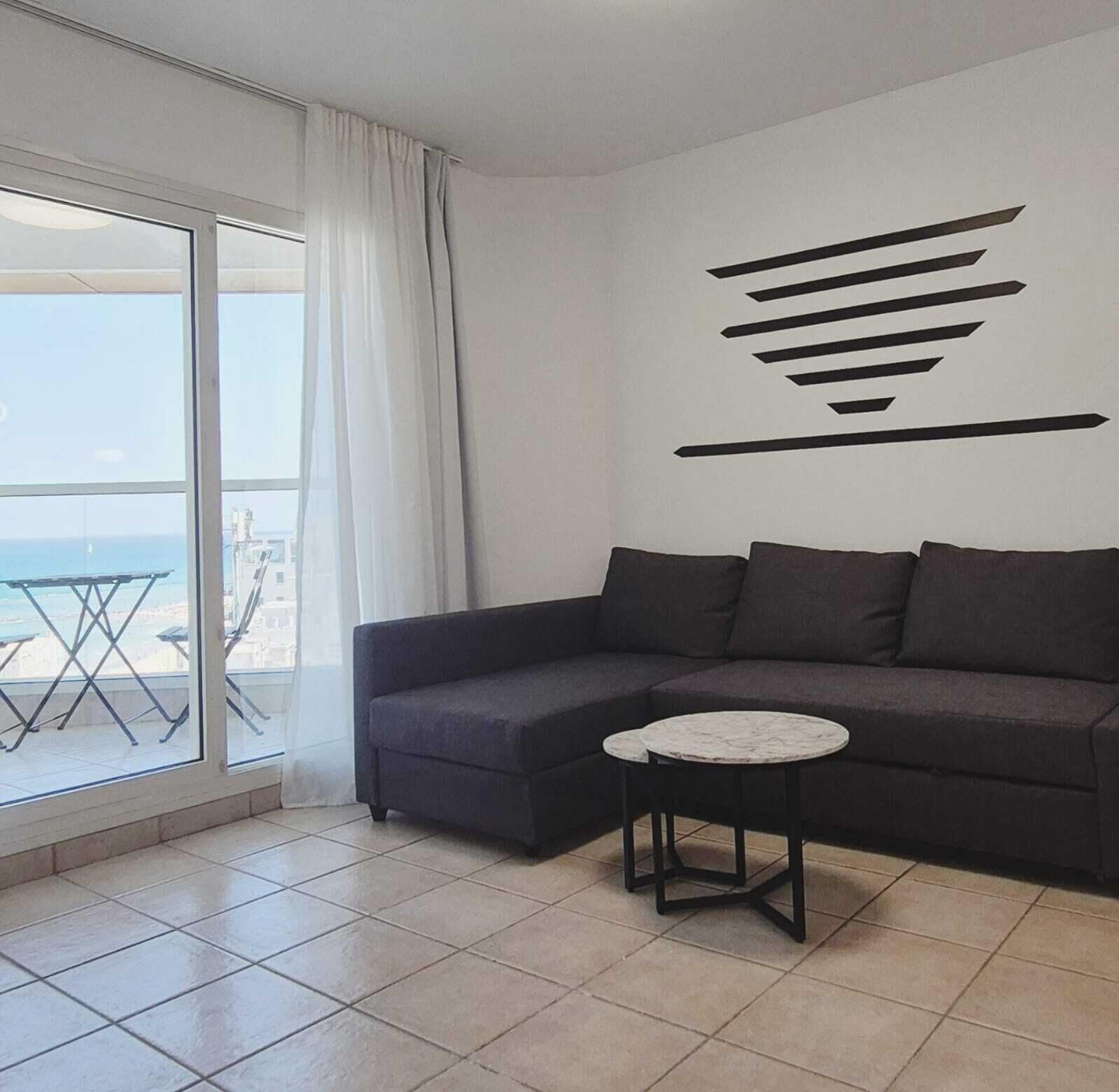 Uma sala de estar moderna com um sofá cinza escuro, uma mesinha redonda e uma janela ampla com vista para o mar.