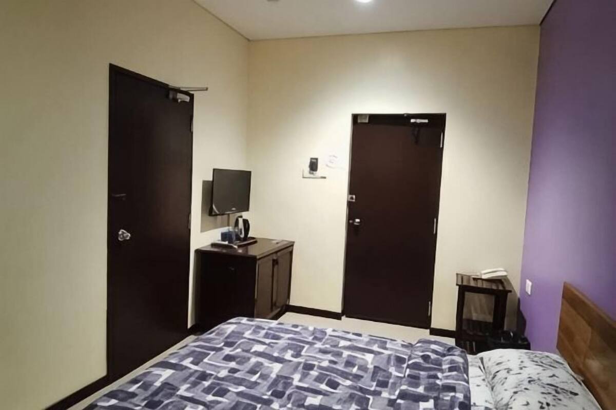 Quarto com cama, televisão em móvel, duas portas e uma pequena mesa com abajur.