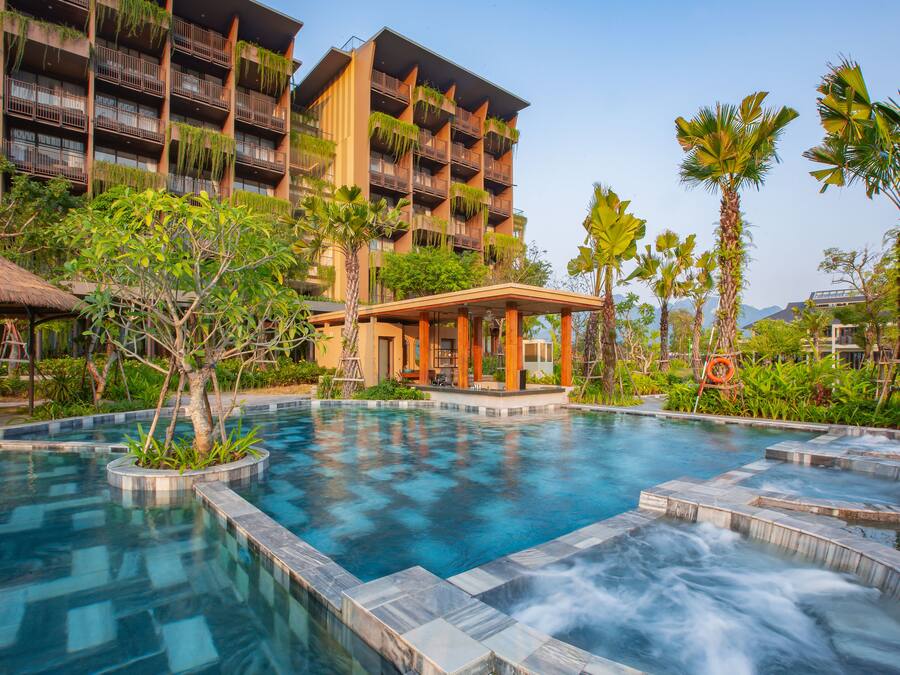 Um hotel com uma grande piscina ao ar livre, várias varandas com plantas e um jardim tropical.