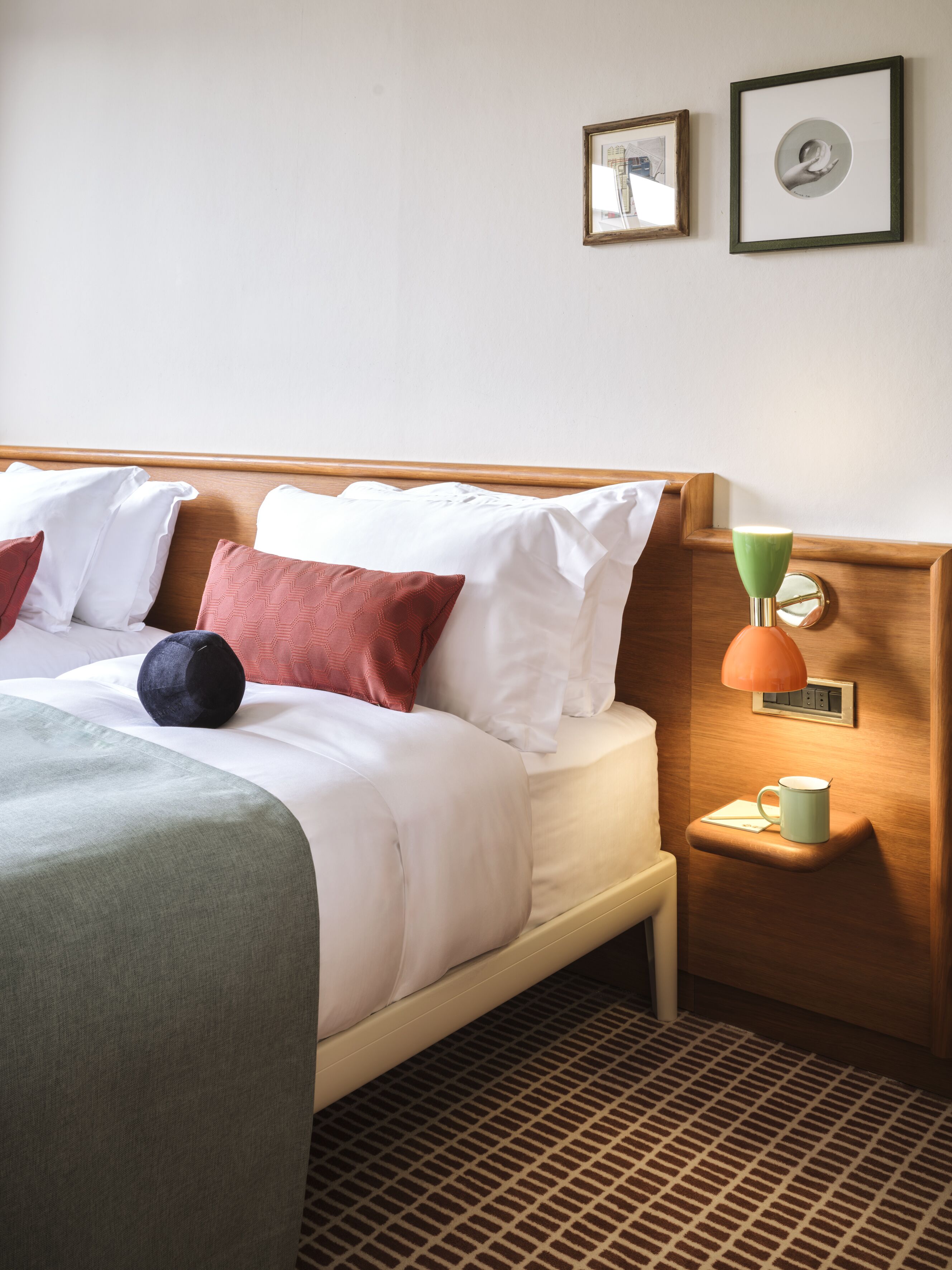 Cama bem arrumada com almofadas brancas e vermelhas, um cobertor verde e cabeceira de madeira. Tem uma mesinha de cabeceira com uma luminária verde e uma caneca. Duas fotos emolduradas estão penduradas na parede acima da cama.