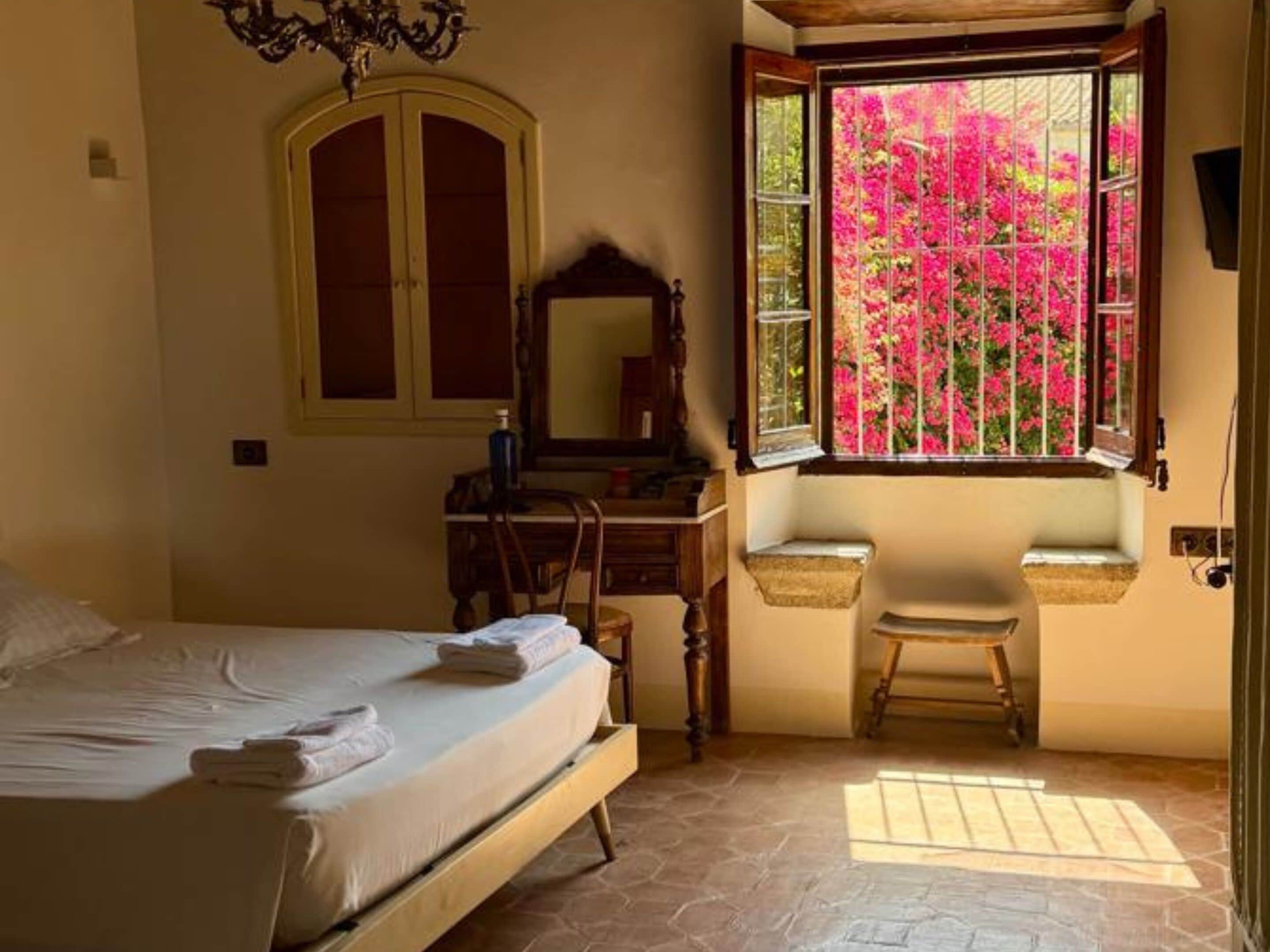 Quarto com cama, escrivaninha, cadeira, janela e vista para flores cor-de-rosa, além de um espelho.