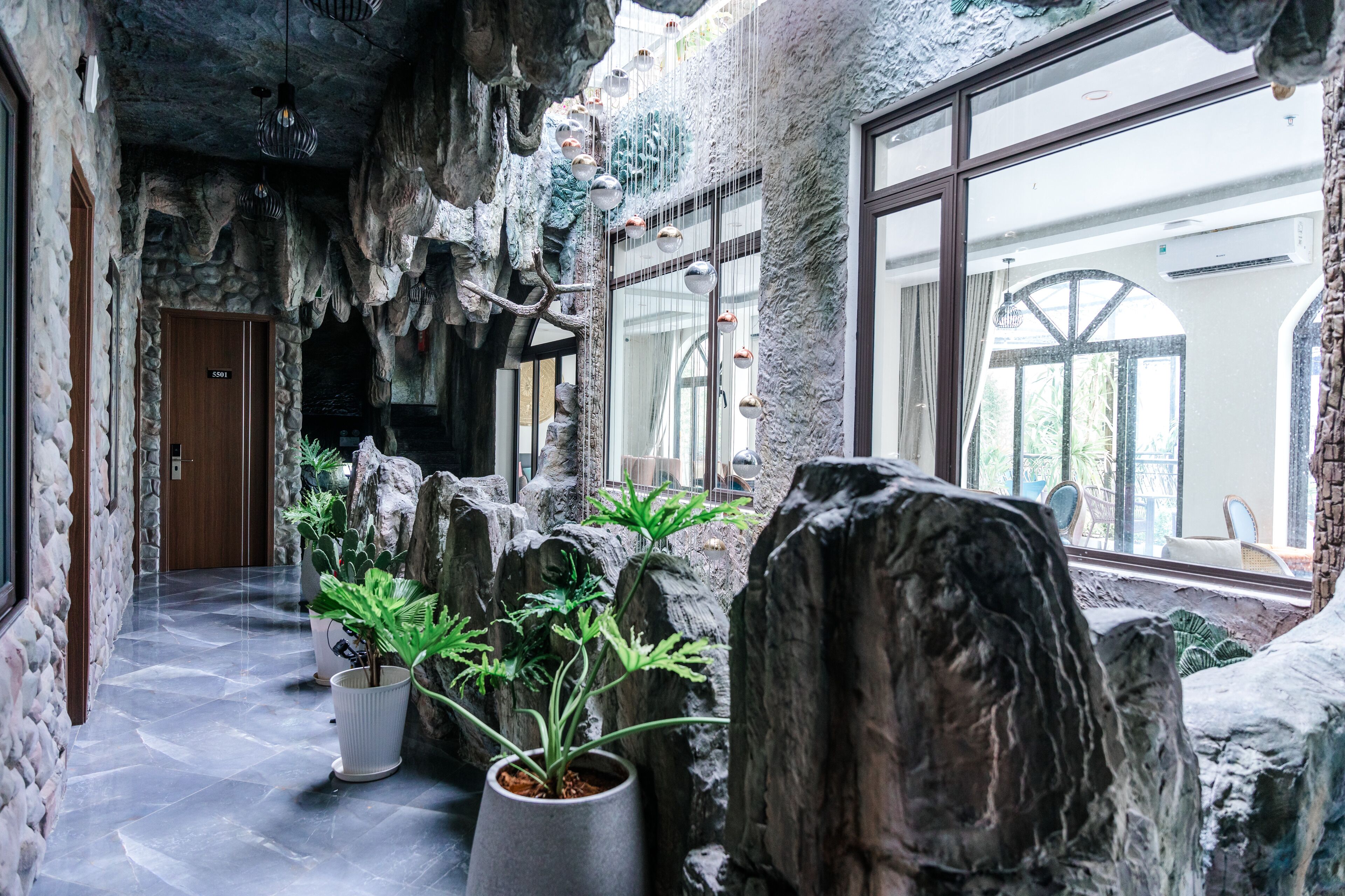 Um corredor de paredes de pedra com uma porta de madeira, plantas em vasos e grandes formações rochosas.