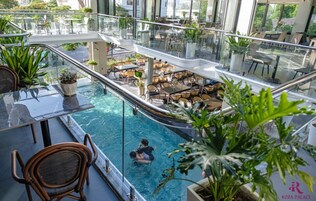 Piscina interior em vários níveis, com guarda-corpos de vidro, rodeada por áreas de descanso com cadeiras, mesas e plantas em vasos.
