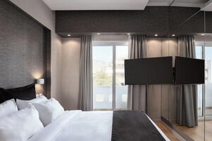 Quarto de hotel moderno com uma cama grande, televisão de tela plana na parede e uma janela ampla com cortinas.