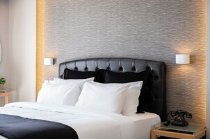 Quarto de hotel moderno com cabeceira estofada, roupa de cama branca e um telefone antigo em uma mesa de cabeceira.