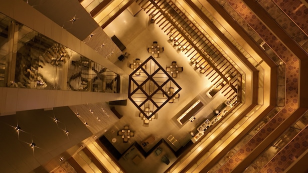 Um átrio amplo, com pé-direito alto, luminária geométrica central, diversas áreas de estar e piso trabalhado.