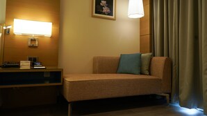 Quarto de hotel moderno com sofá, mesa de cabeceira com abajur, relógio e um quadro na parede.
